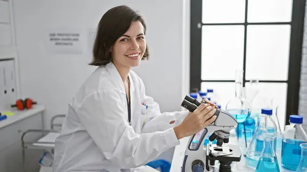 Smiling young hispanic woman scientist joyfully working with microscope ...