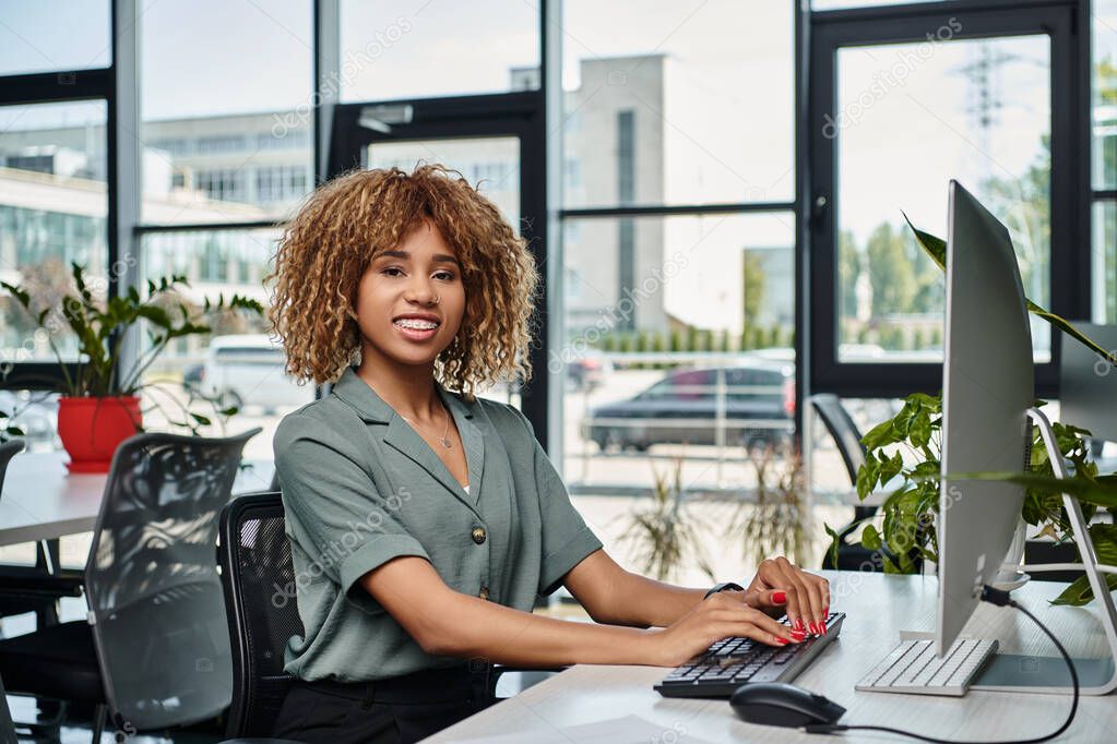 feliz joven mujer de negocios afroamericana sentada en el escritorio frente al monitor de la ...
