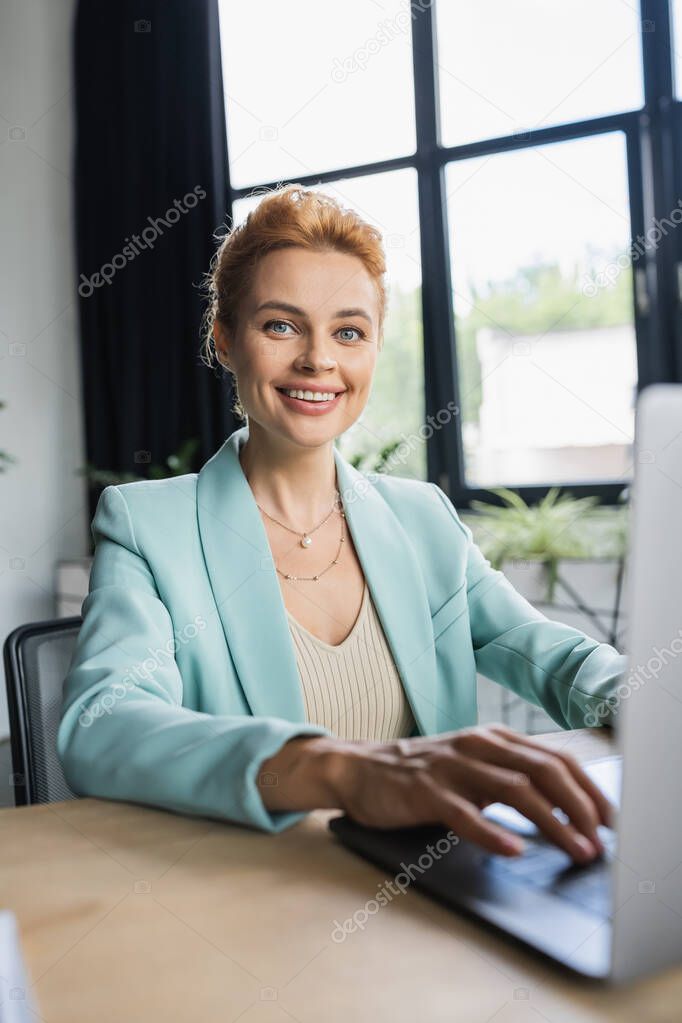 exitosa mujer de negocios en blazer elegante escribiendo en el ordenador portátil y sonriendo a ...