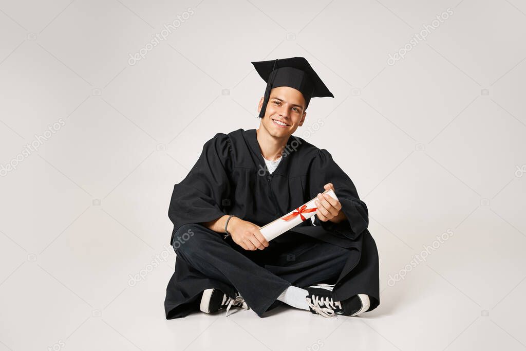 hombre guapo en vestido de graduado y gorra sentado y sosteniendo el ...