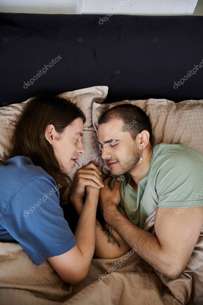 Una joven pareja gay yace en la cama, tomados de la mano y mirándose cariñosamente. 2025