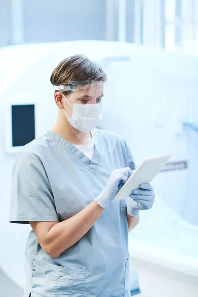 Busy young MRI tech in safety face shield and gloves using tablet while ...
