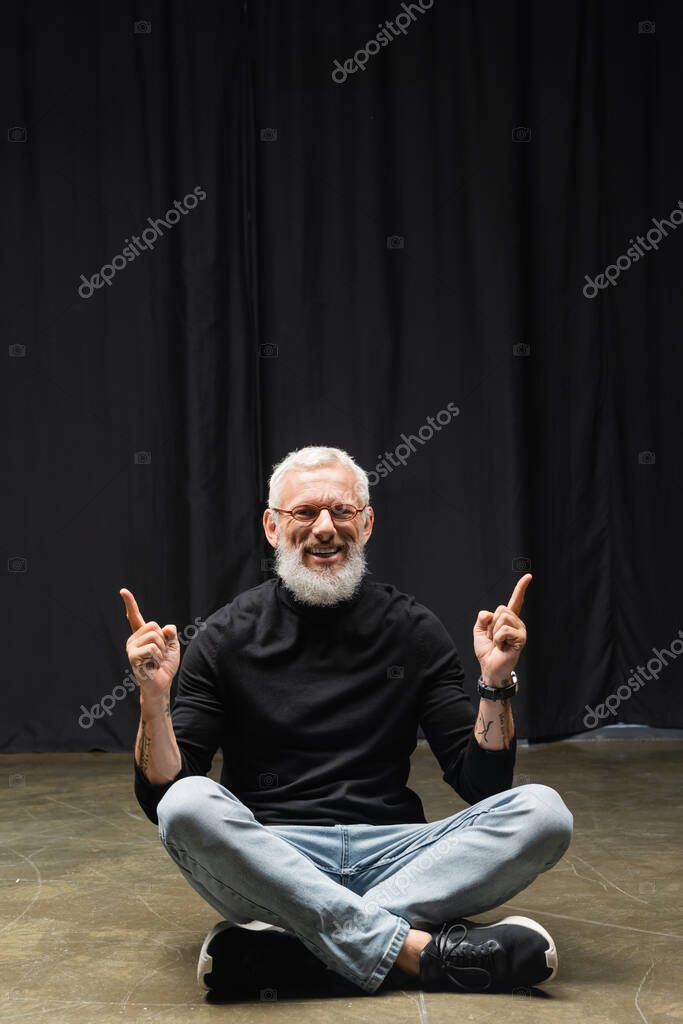cheerful tattooed actor pointing up with fingers while sitting on ...