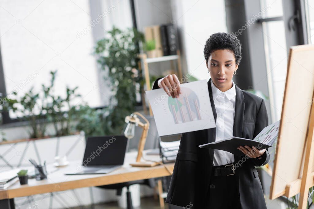 joven mujer de negocios afroamericana sosteniendo carpeta y gráficos cerca del tablero de notas ...