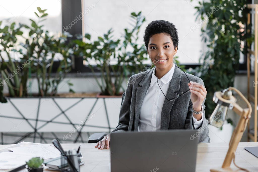 alegre mujer de negocios afroamericana en traje gris con gafas y mirando a la cámara cerca de la ...