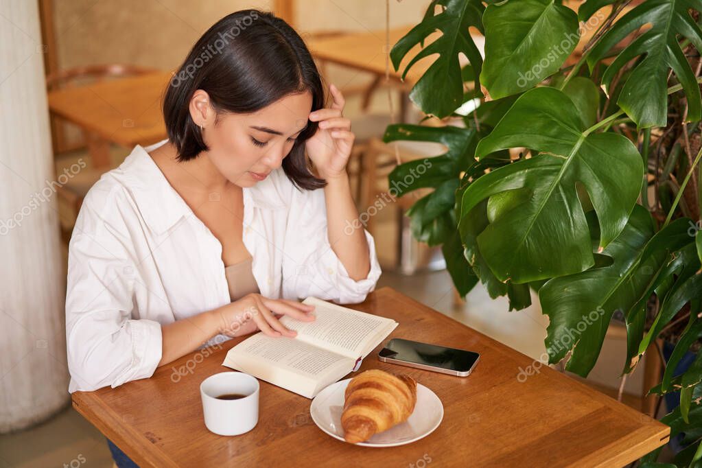 Hermosa joven asiática sentada en la cafetería con un libro, comiendo ...