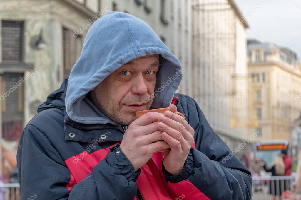 A homeless man 45-50 years old in a jacket and hood with a mug in his ...