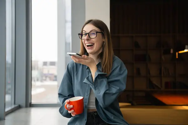 Cheerful young woman in glasses recording voice message in library ...